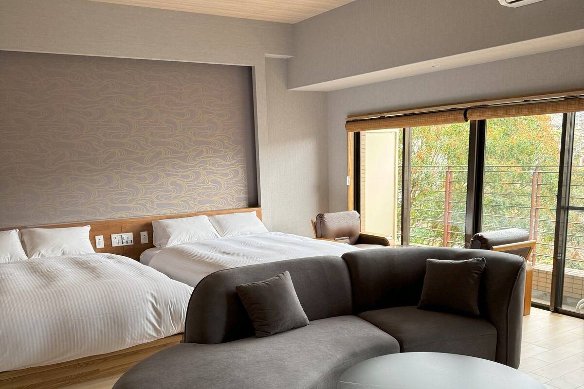 Quarto de hotel moderno com uma cama grande, um sofá, uma mesa redonda e vista para árvores através de janelas amplas.