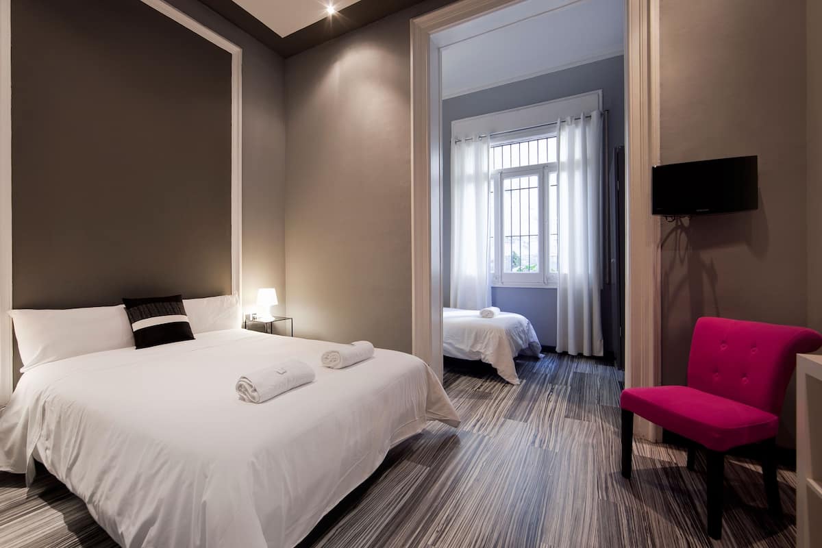 Quarto de hotel com uma cama grande, uma cama menor, uma TV fixada na parede e uma janela iluminada com cortinas.