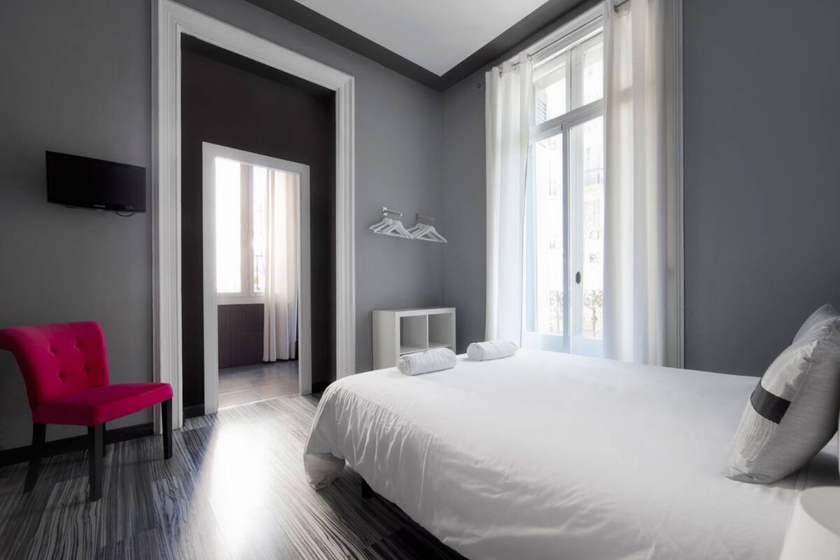 Quarto de hotel moderno com uma cama grande, uma cadeira vermelha, uma TV na parede e vista para a cidade pelas janelas.