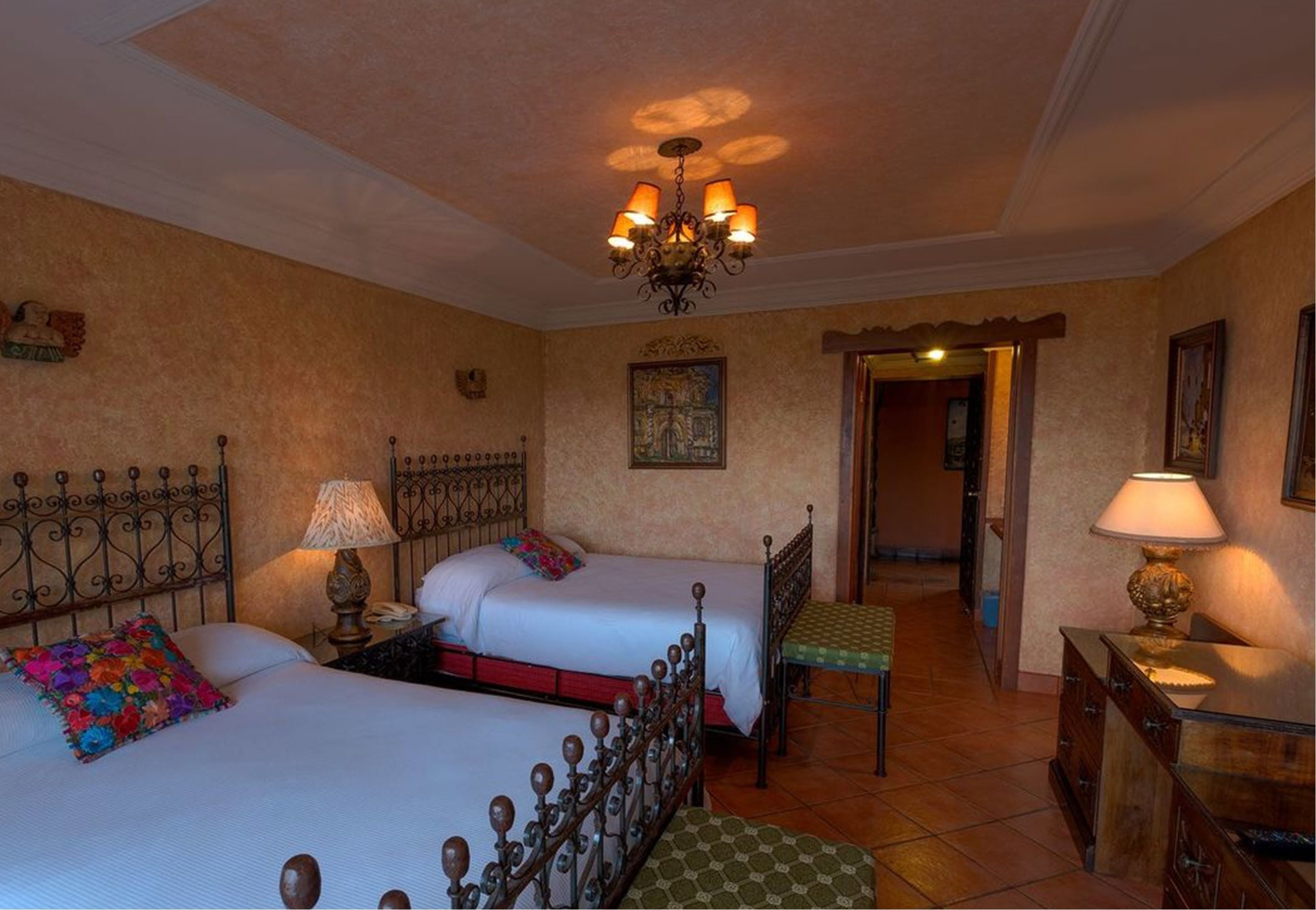 Quarto de hotel com duas camas, um candelabro, uma escrivaninha e uma cadeira.