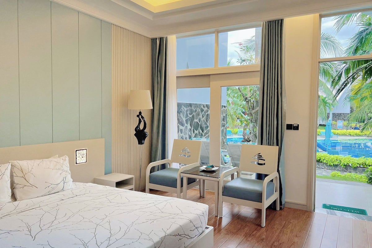 Quarto de hotel com uma cama, duas cadeiras, uma mesa e uma janela grande com vista para uma piscina e vegetação.