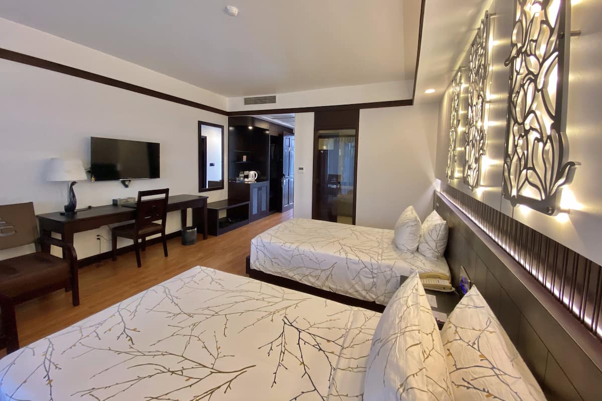 Quarto de hotel com duas camas, uma escrivaninha, uma cadeira, uma TV e um painel decorativo na parede.