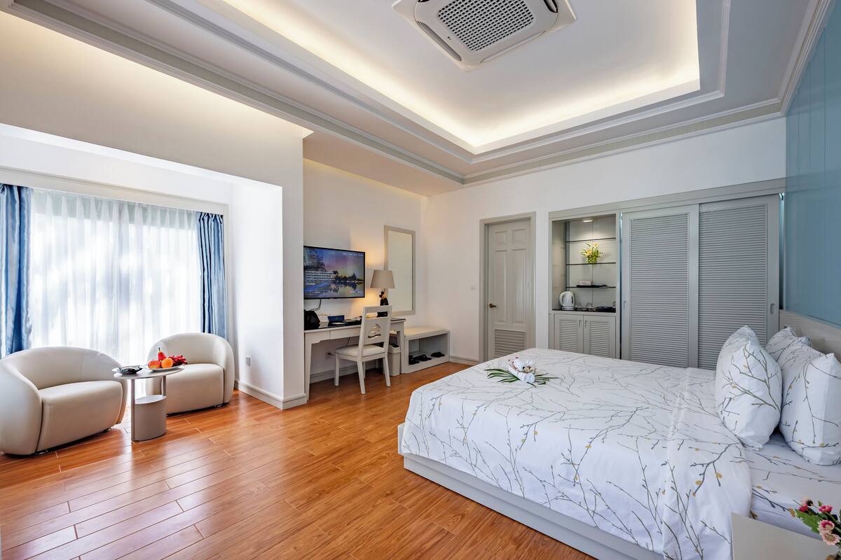 Quarto de hotel moderno com piso de madeira, uma cama com colcha floral branca, uma escrivaninha com cadeira, um sofá e uma televisão.