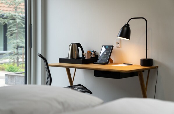 Quarto de hotel minimalista com escrivaninha de madeira, luminária de mesa preta, um laptop, máquina de café e um tabuleiro preto com diversos objetos.