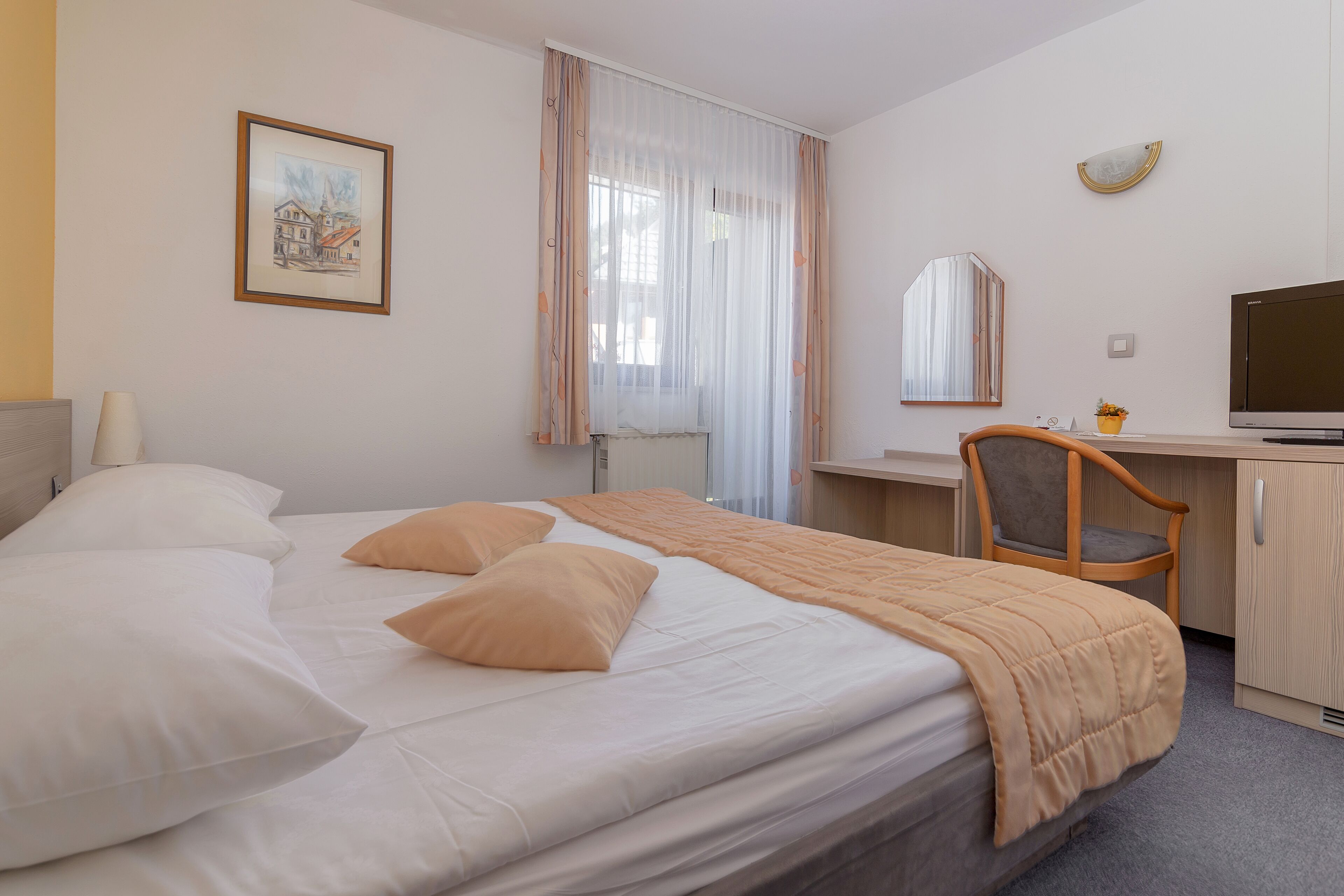 Quarto de hotel com cama, escrivaninha, cadeira, televisão e um quadro na parede.