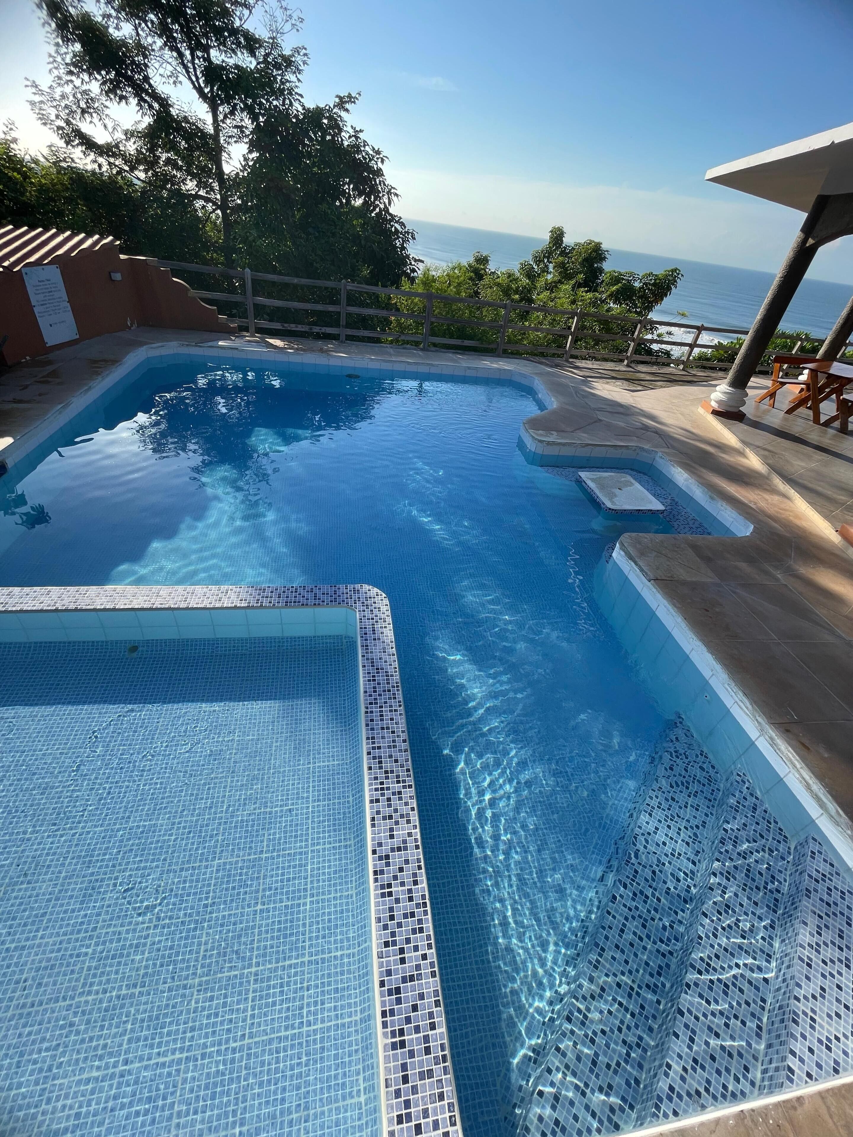 Piscina com água cristalina, rodeada por um deck ladrilhado e uma cerca de madeira.