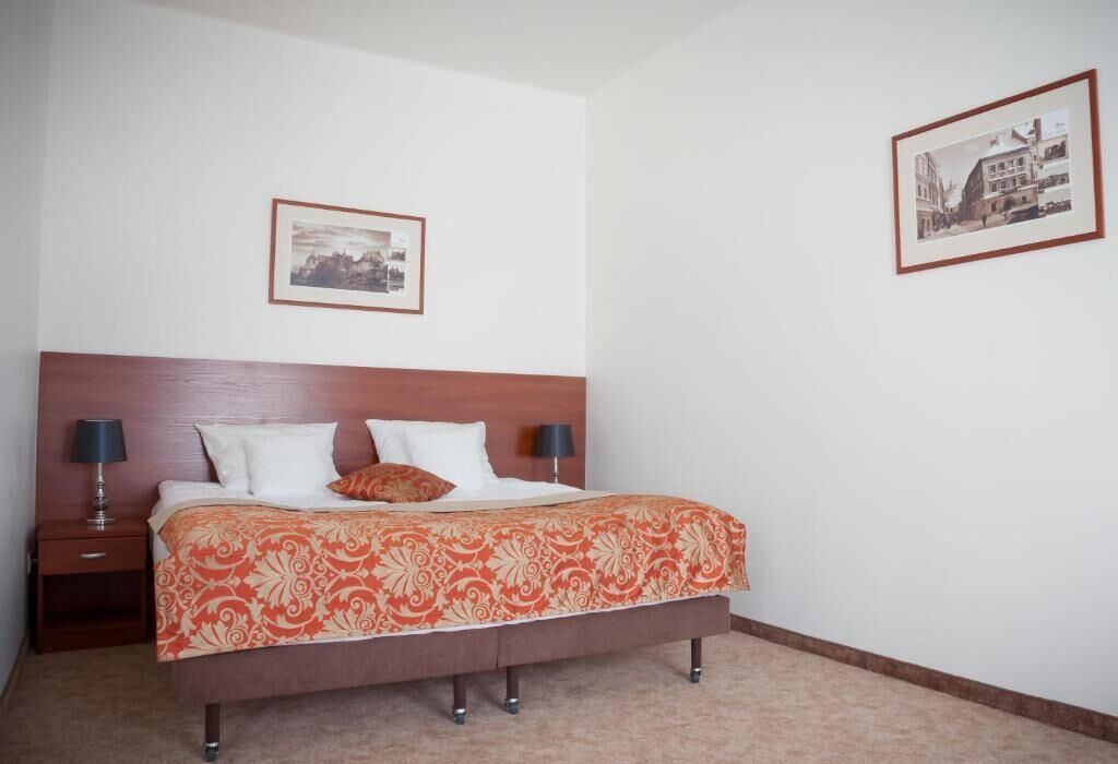 Quarto de hotel com cabeceira de madeira, cama com colcha laranja estampada, duas luminárias de cabeceira e duas fotos emolduradas nas paredes.