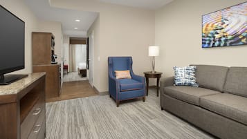 Quarto de hotel com televisão, uma poltrona azul com almofada estampada, sofá cinza com travesseiro azul e branco, mesa lateral pequena com luminária e uma grande pintura abstrata na parede.