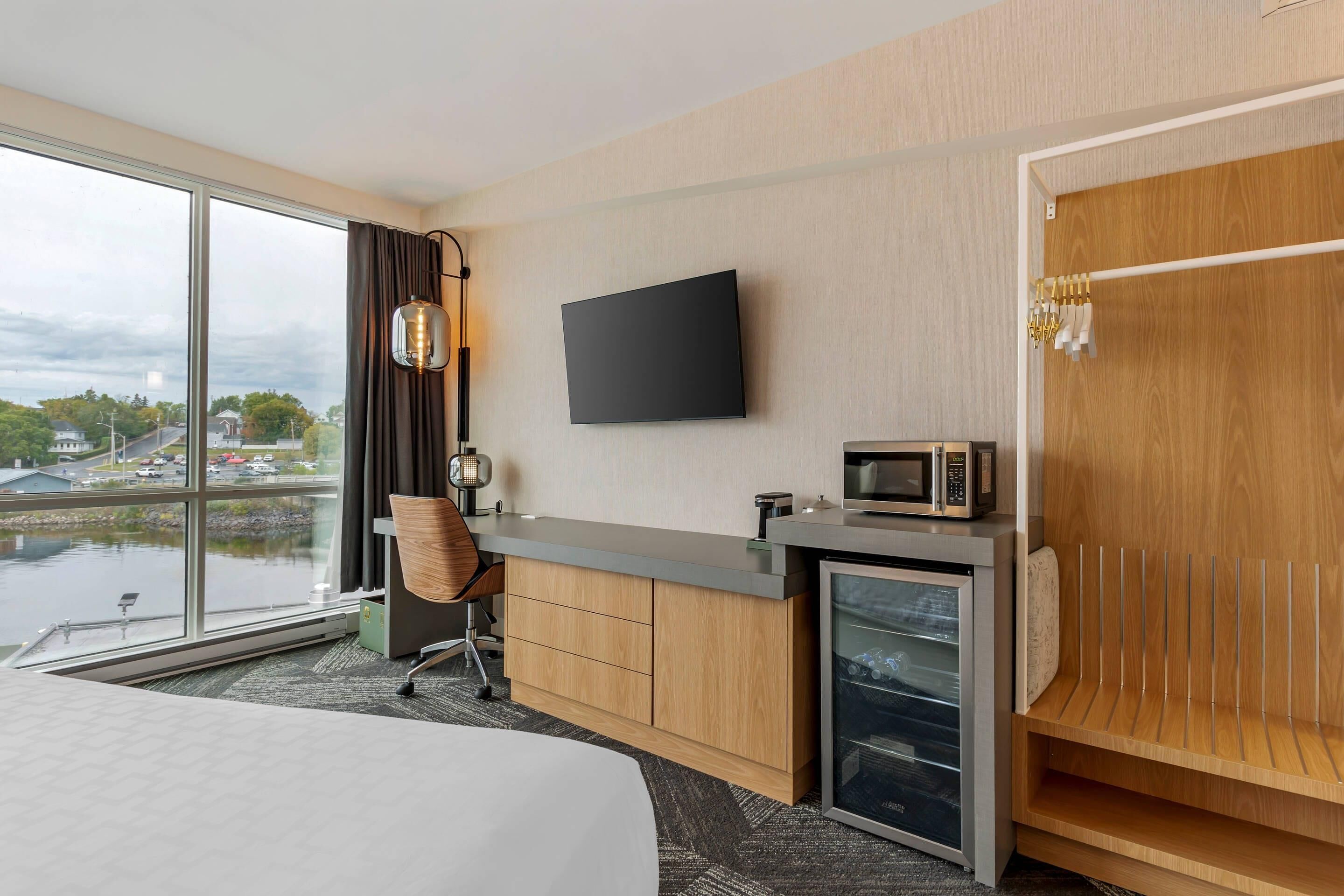 Quarto de hotel com uma cama grande, escrivaninha com micro-ondas, televisão e vista para um corpo de água.