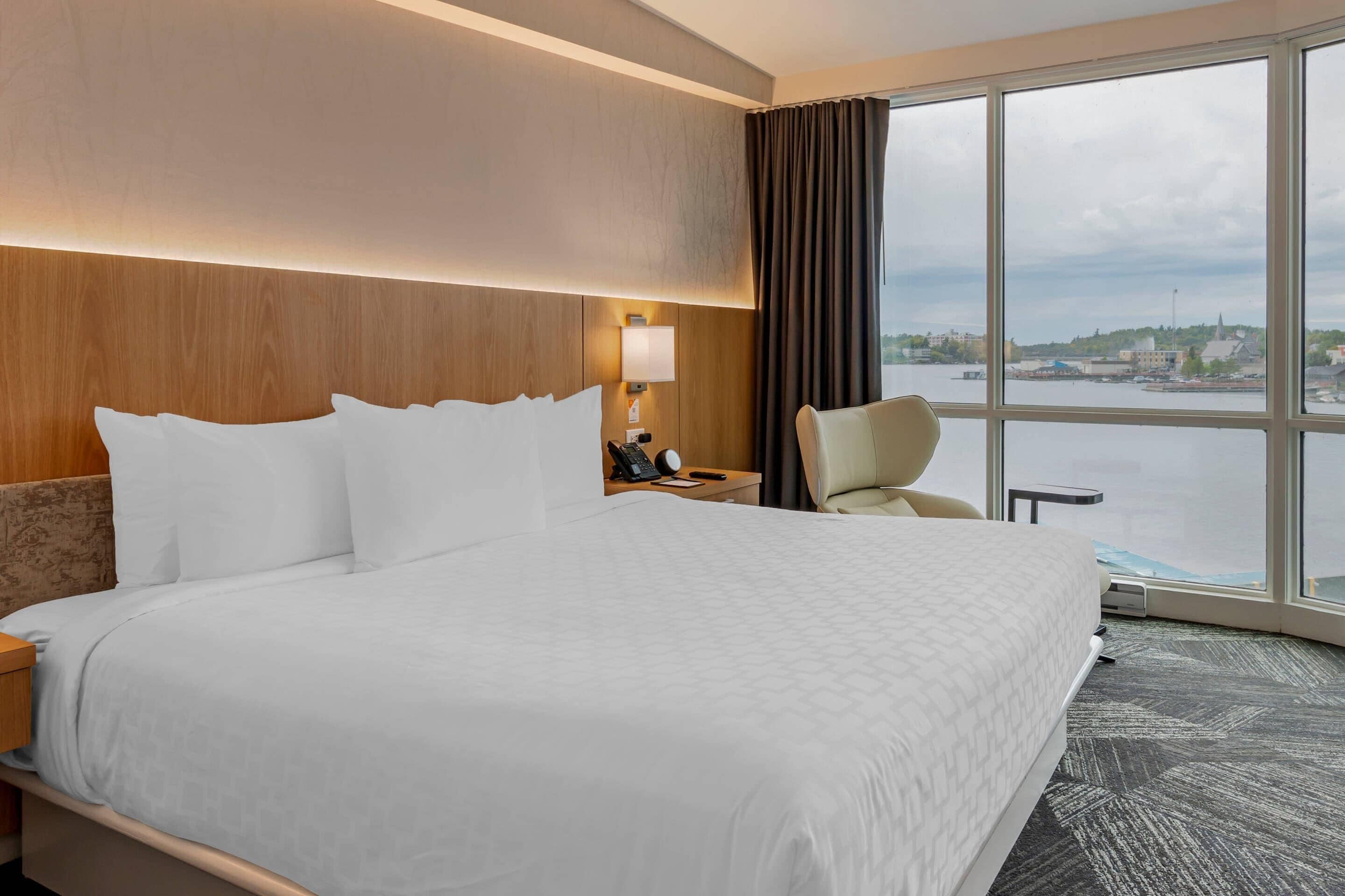Quarto de hotel com uma cama grande, mesa de cabeceira com abajur, uma cadeira e vista para um corpo de água através de janelas amplas.