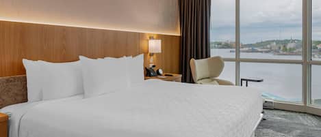 Quarto de hotel com uma cama grande, mesa de cabeceira com abajur, uma cadeira e vista para um corpo de água através de janelas amplas.
