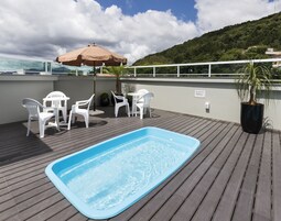 Uma área de piscina no terraço com piscina inflável azul, cadeiras de plástico branco, um guarda-sol bege e um vaso de planta.