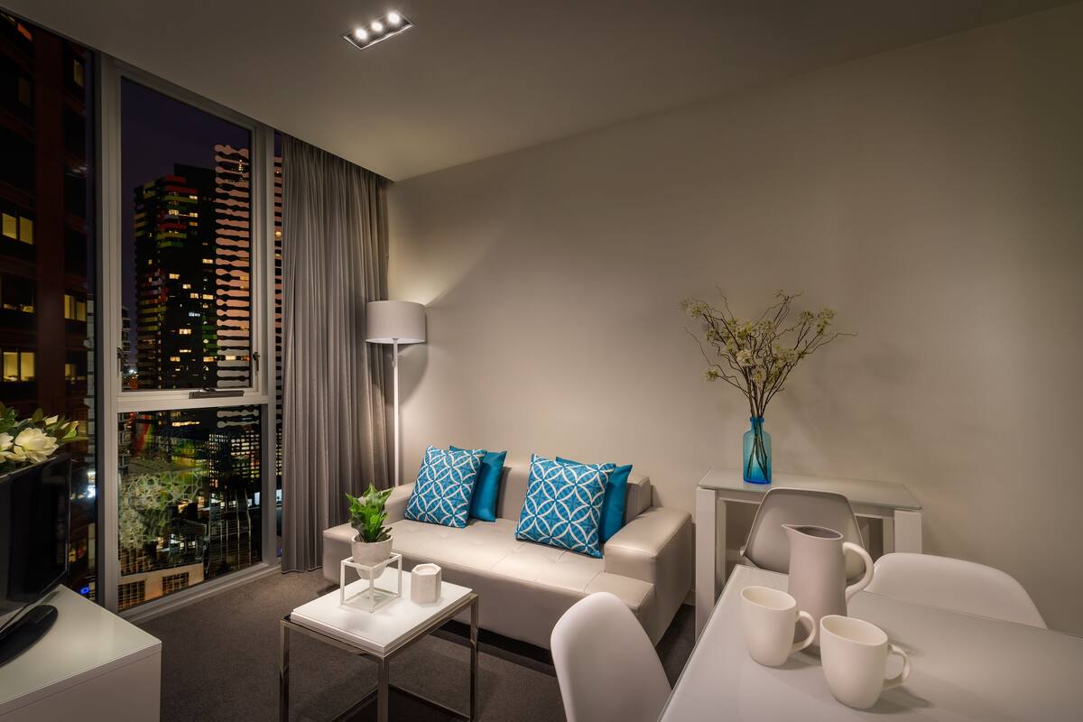 Quarto de hotel moderno com uma grande janela, um sofá com almofadas estampadas, uma mesa pequena com uma planta e um vaso com flores.