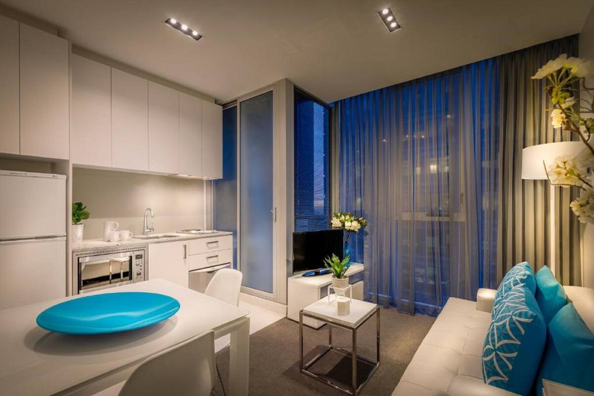 Quarto de hotel moderno com cozinha, mesa de jantar com um prato azul, sofá com almofadas azuis e uma televisão.