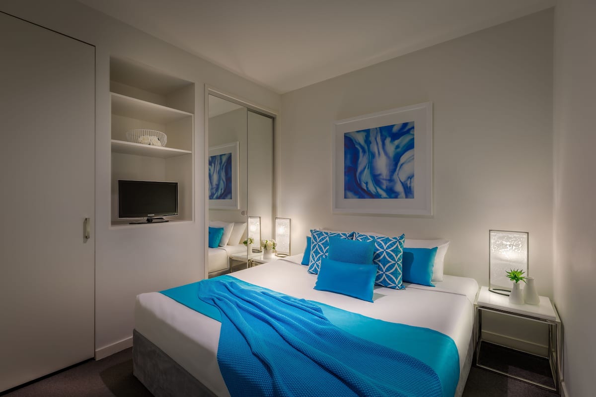 Quarto de hotel moderno com uma cama grande, televisão, espelho e mesa de cabeceira com abajur.