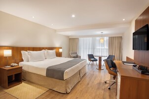 Quarto de hotel com uma cama grande, mesas de cabeceira, escrivaninha com cadeiras, televisão e janela com cortinas.