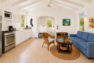 Cozinha moderna com armários brancos, fogão e micro-ondas. Na sala de estar, há uma cadeira de vime e um sofá azul, com um tapete tecido no chão.