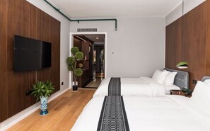 Quarto de hotel moderno com uma televisão de tela grande, cama com roupa de cama branca, mesinha de cabeceira pequena e um vaso de plantas.