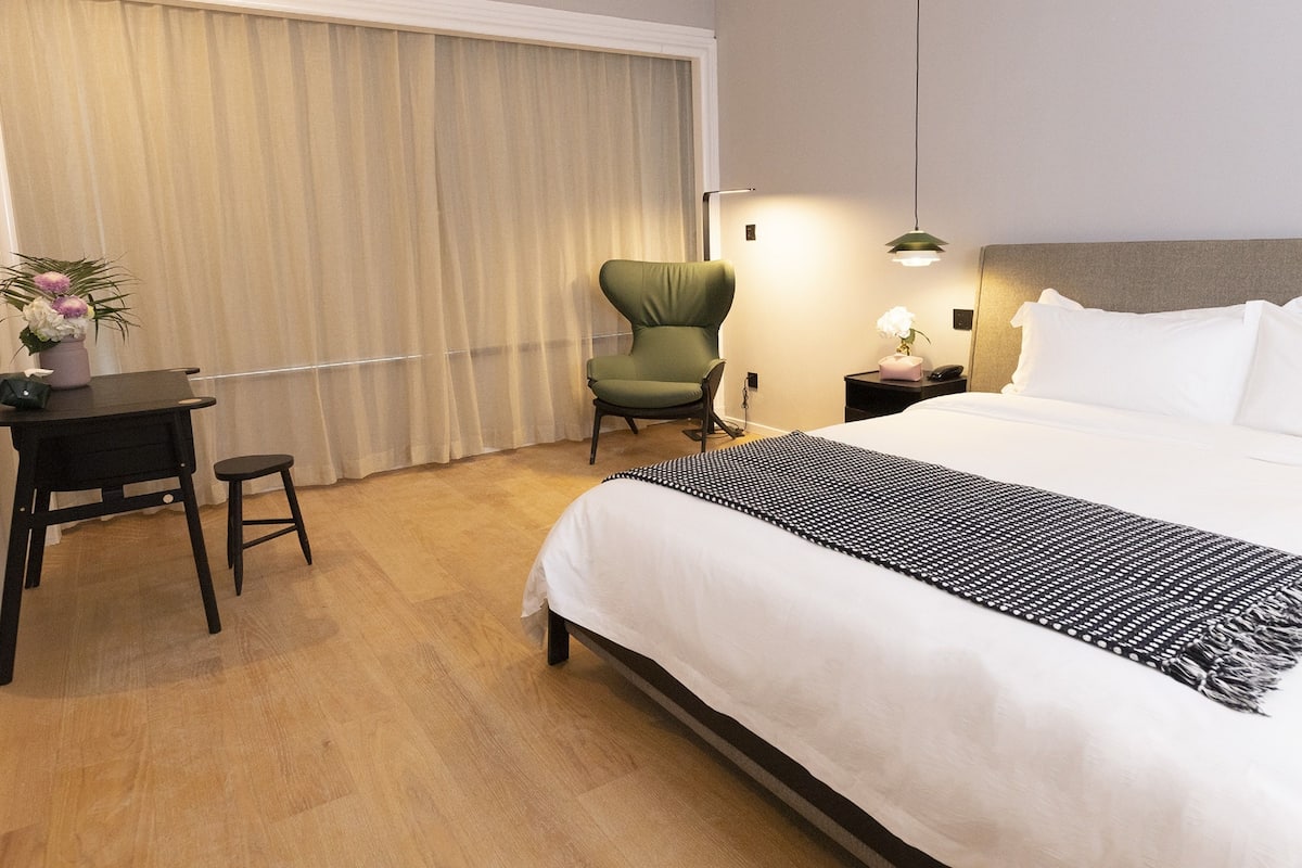 Quarto de hotel com cama, escrivaninha, cadeira, mesa de cabeceira e abajur.
