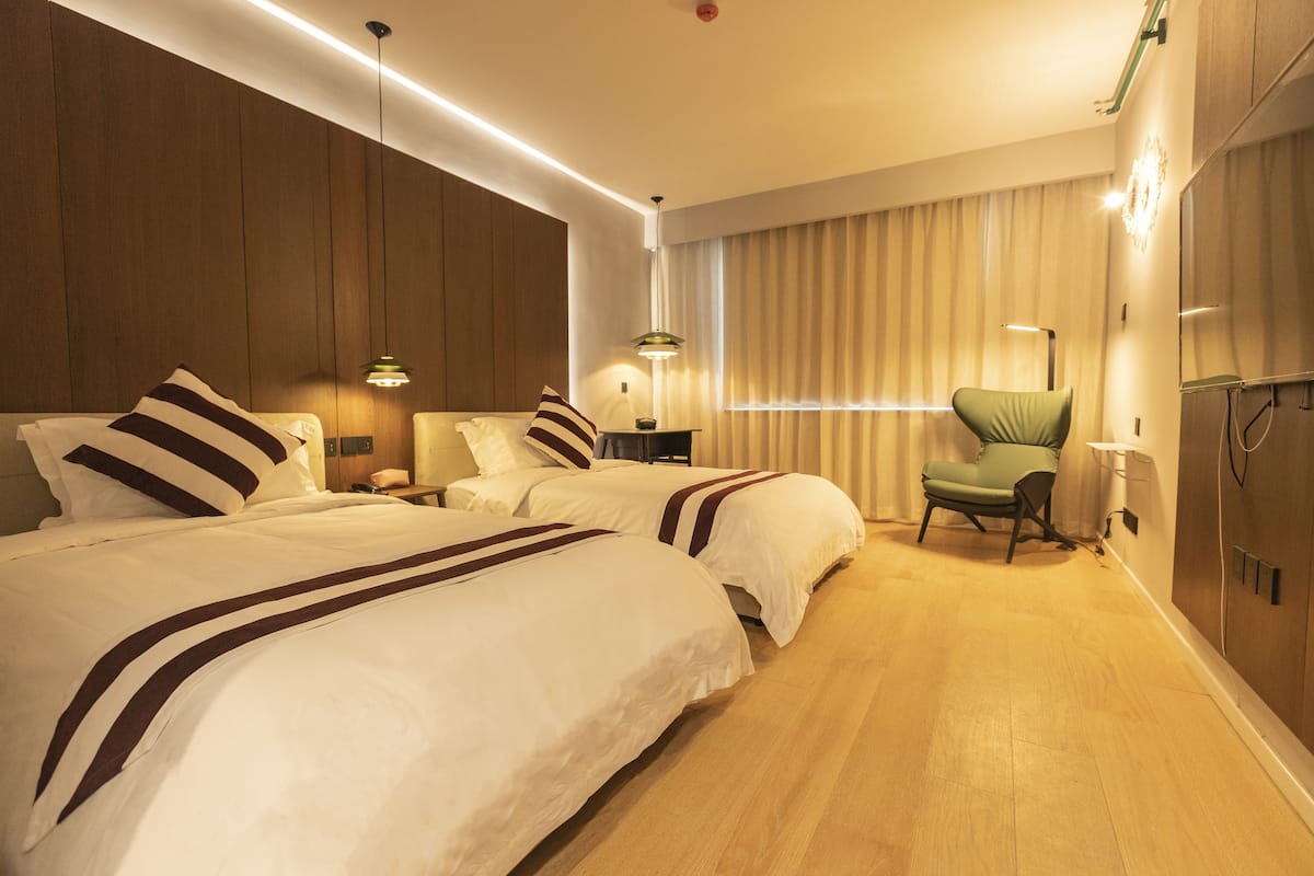 Quarto de hotel com duas camas, painéis de madeira nas paredes, uma cadeira verde e uma janela grande com cortinas.