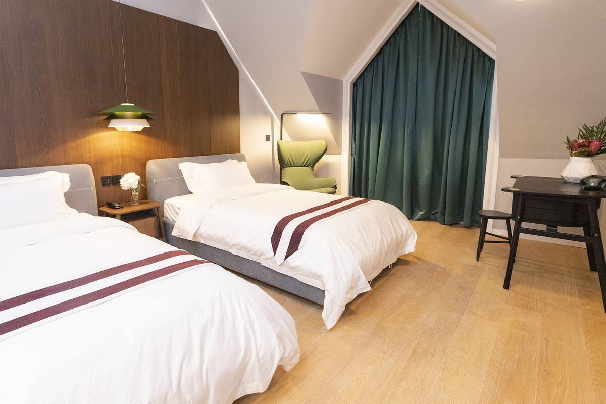 Quarto de hotel com duas camas, uma escrivaninha, uma cadeira e uma janela grande com cortinas.