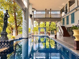 Uma área de piscina com uma estátua, uma galeria coberta e uma escadaria que leva a um andar superior.
