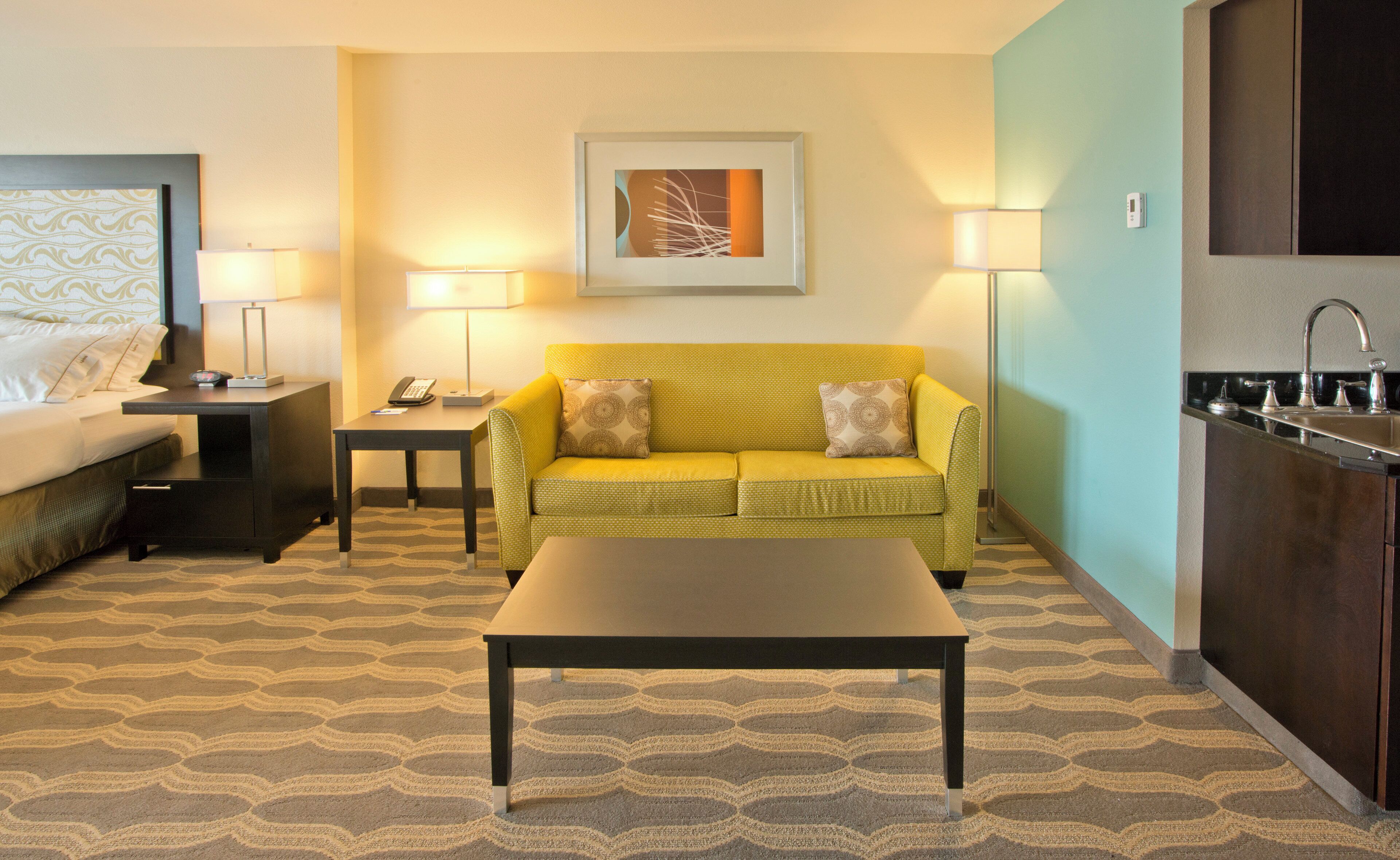 Quarto de hotel com cama, sofá amarelo, mesa pequena, mesa de cabeceira, luminária, quadro e cozinha compacta.