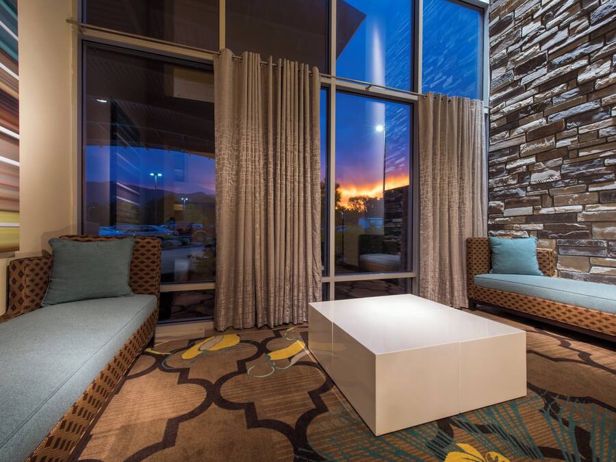 Quarto de hotel moderno com uma grande janela, um tapete estampado, uma mesa de centro branca e uma parede de pedra.