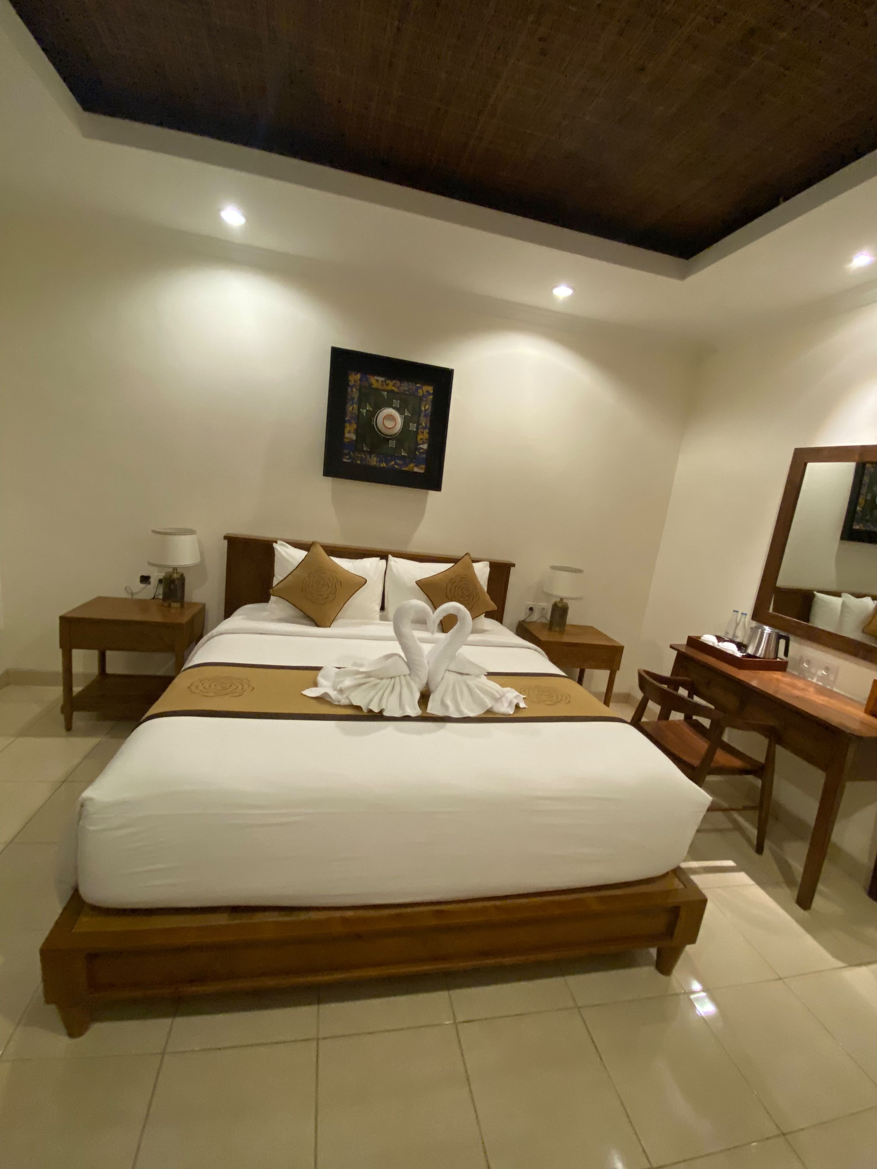 Quarto de hotel com cama grande, cabeceira de madeira, escrivaninha com cadeira, mesa de cabeceira, relógio de parede e espelho.