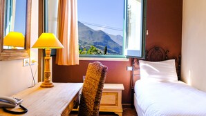 Quarto de hotel com uma cama, uma cadeira, uma mesa de trabalho com abajur e vista para montanhas pela janela.