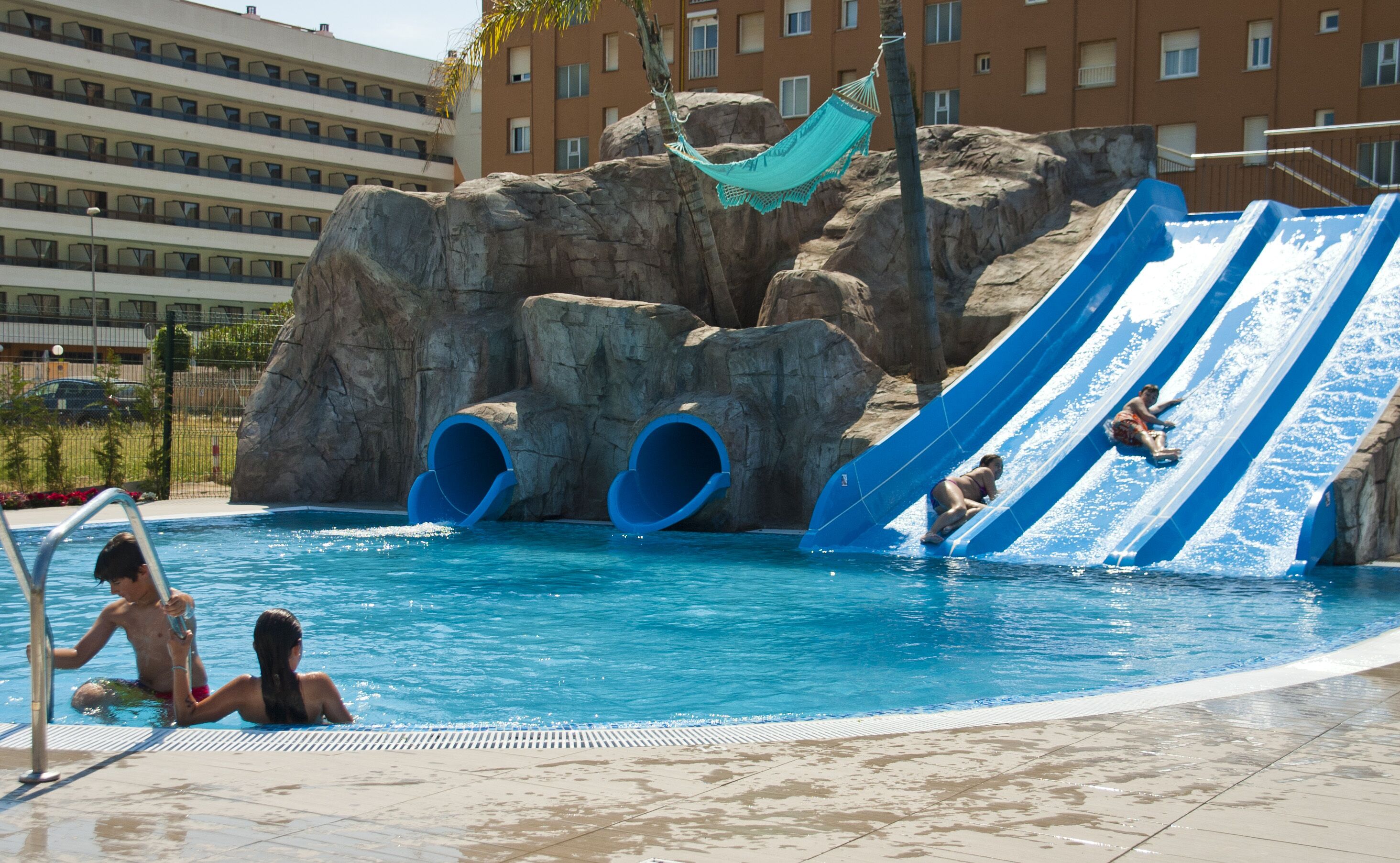 Piscina com escorregadores, uma formação rochosa e um prédio ao fundo.