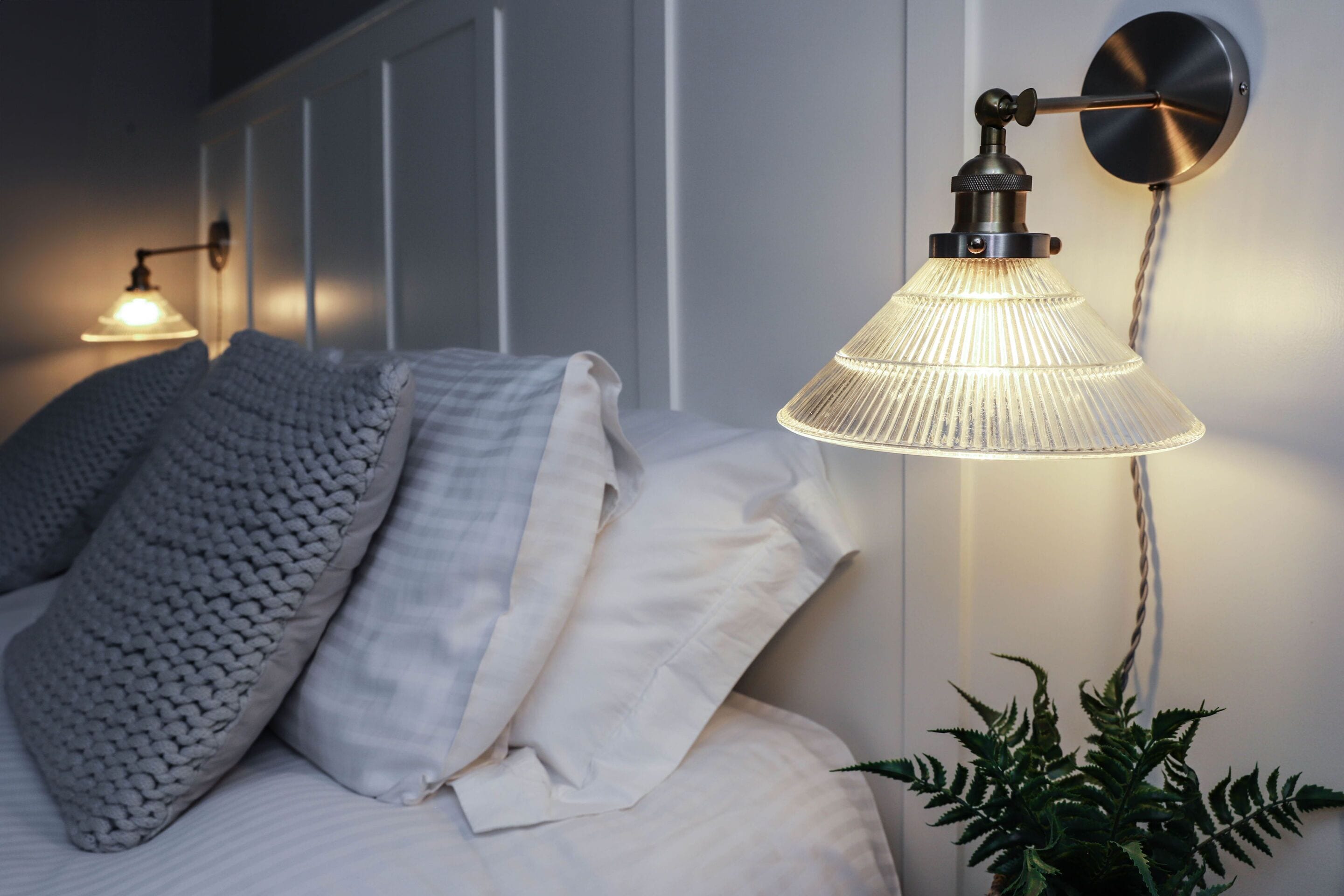 Uma cama bem arrumada com manta branca e cinza, uma luminária na parede e um vaso com planta.