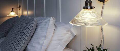 Uma cama bem arrumada com manta branca e cinza, uma luminária na parede e um vaso com planta.