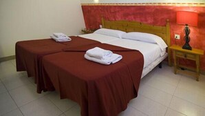 Quarto de hotel com duas camas de solteiro, cada uma com um edredom vermelho e uma toalha branca dobrada. Há uma mesa de cabeceira de madeira com uma luminária à direita das camas.