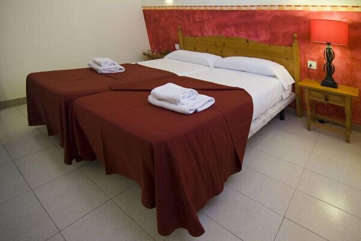 Quarto de hotel com duas camas de solteiro, cada uma com um edredom vermelho e uma toalha branca dobrada. Há uma mesa de cabeceira de madeira com uma luminária à direita das camas.