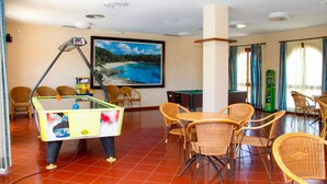 Quarto com mesa de bilhar, cadeiras de vime, mesa de jantar e uma grande pintura de uma paisagem praiana.