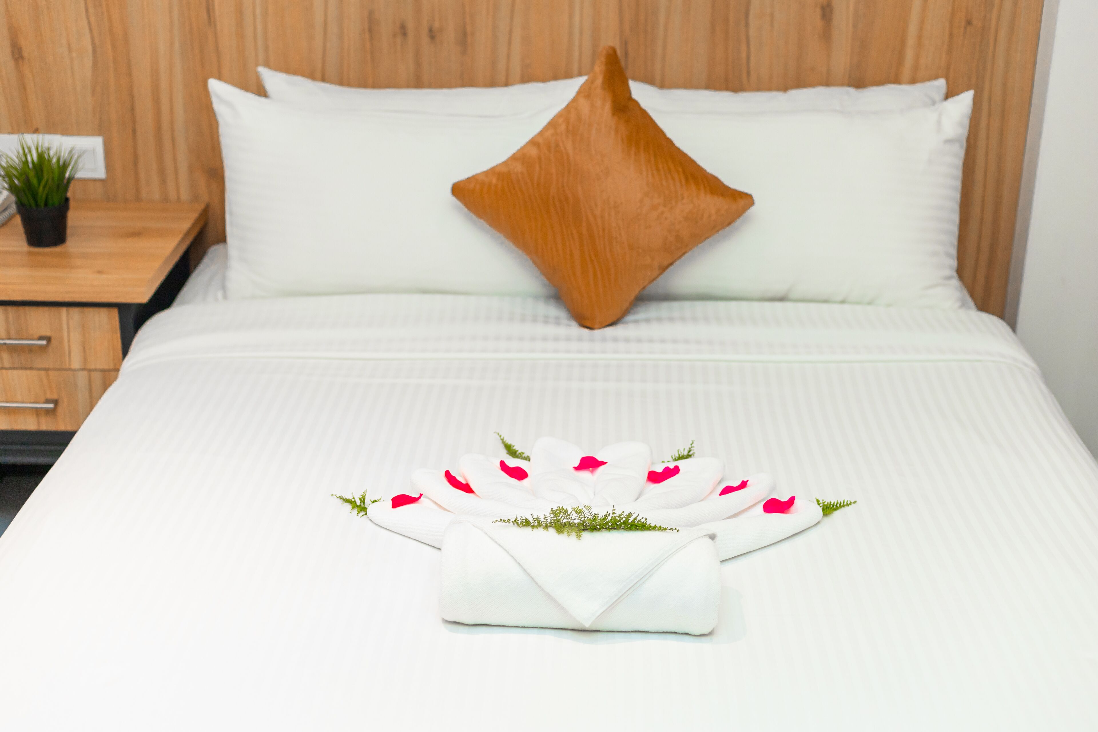 Uma cama bem arrumada com colcha branca, travesseiro marrom decorativo e um arranjo de toalhas brancas e folhagem verde.