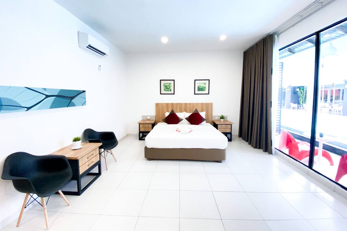 Quarto de hotel moderno com uma cama grande, uma escrivaninha com cadeira, uma pequena mesa com planta e uma janela ampla com vista para o exterior.