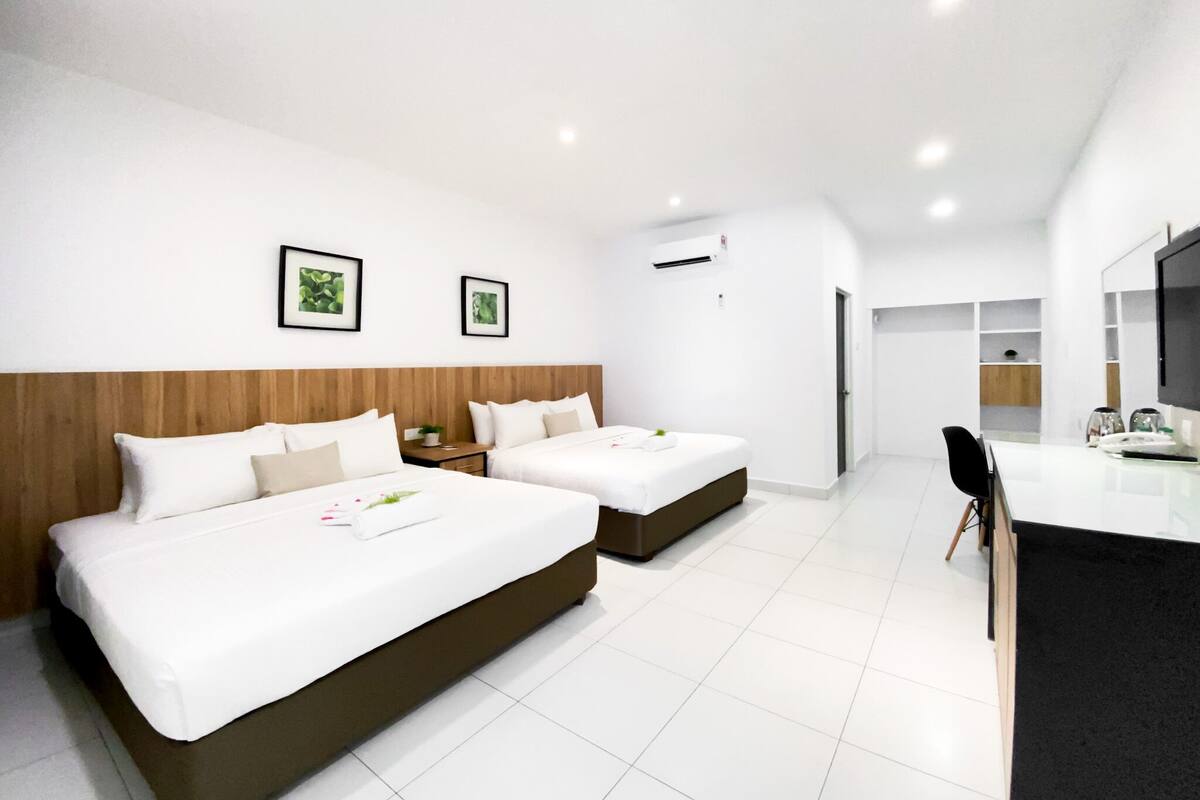 Quarto de hotel com duas camas, cabeceira de madeira, paredes brancas e uma área de cozinha compacta.