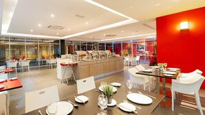 Restaurante moderno com área de buffet central, diversas mesas de jantar e uma parede de destaque vermelha vibrante.