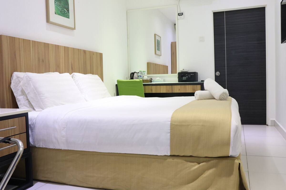 Quarto de hotel com cama, mesa de cabeceira, espelho, cômoda e porta.