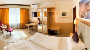 Quarto de hotel com cama, escrivaninha, cadeira, televisão e um quadro na parede.