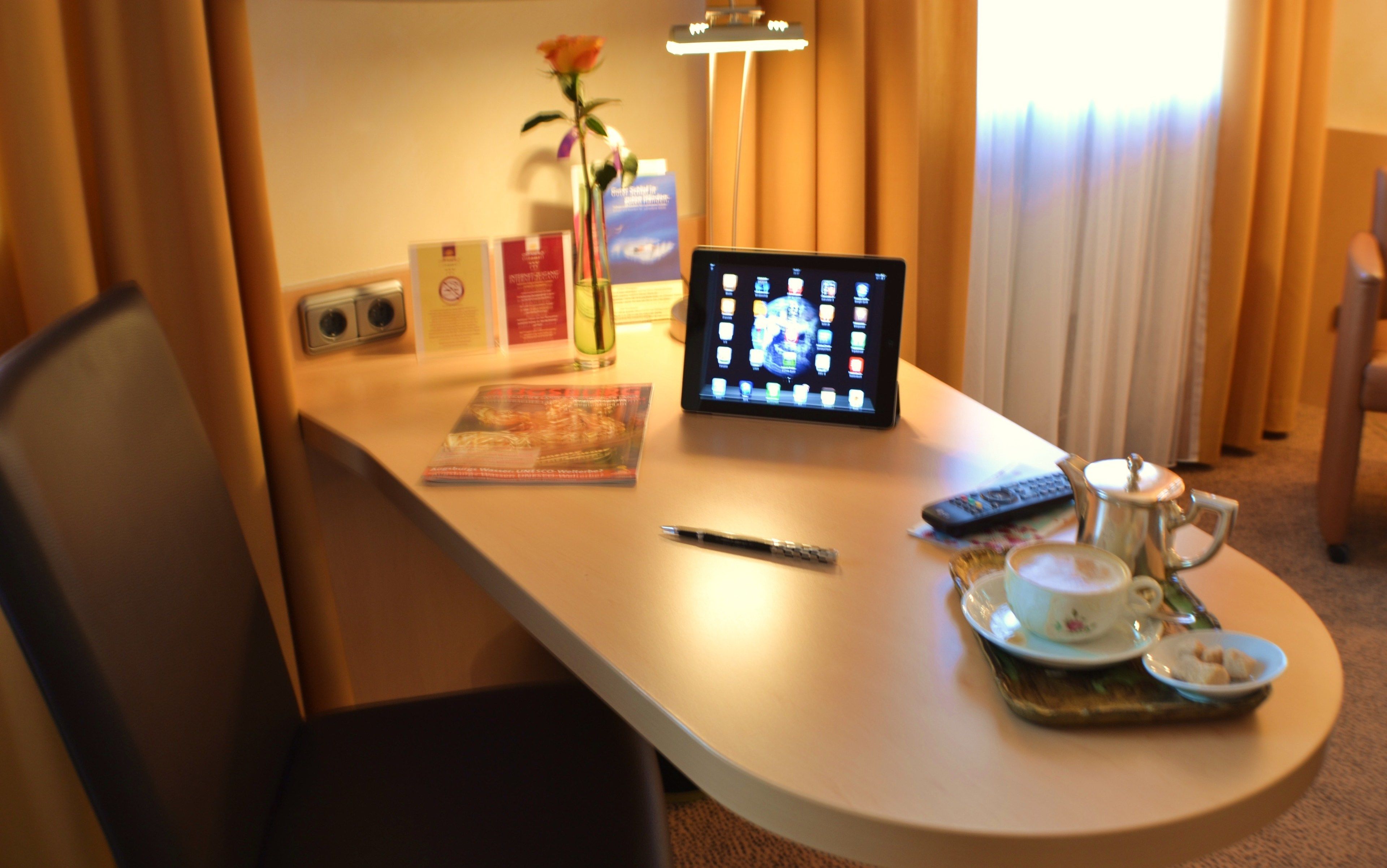 Quarto de hotel com escrivaninha, tablet, controle remoto, caneta, xícara de café e um vaso de flores.
