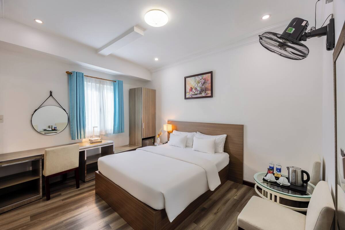 Quarto de hotel com cama, escrivaninha, cadeira, mesa de cabeceira, abajur e ventoinha fixada na parede.