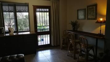 Um quarto com iluminação suave, área de cozinha, mesa de jantar com cadeiras, janela com persianas e porta com vista para o exterior.