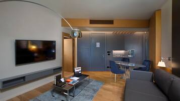 Uma sala de estar moderna com televisão de tela plana, mesa de centro e sofá. Há uma área de jantar com mesa e cadeiras, e uma cozinha ao fundo.
