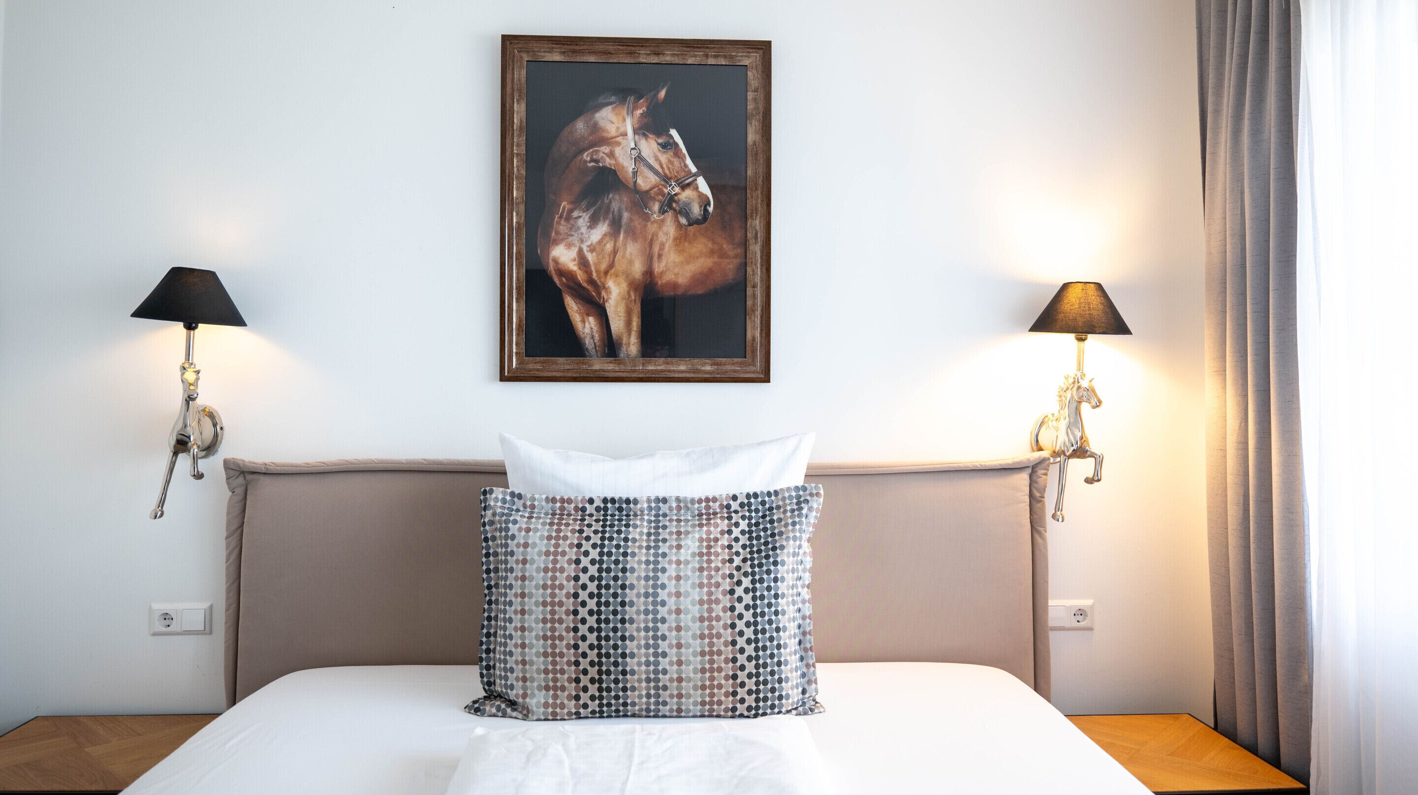 Quarto de hotel com uma cama, almofada estampada, pintura de um cavalo e duas luminárias fixadas na parede.