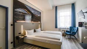 Quarto de hotel com uma cama grande, uma escrivaninha com telefone, uma cadeira, televisão e um quadro de uma ponte.
