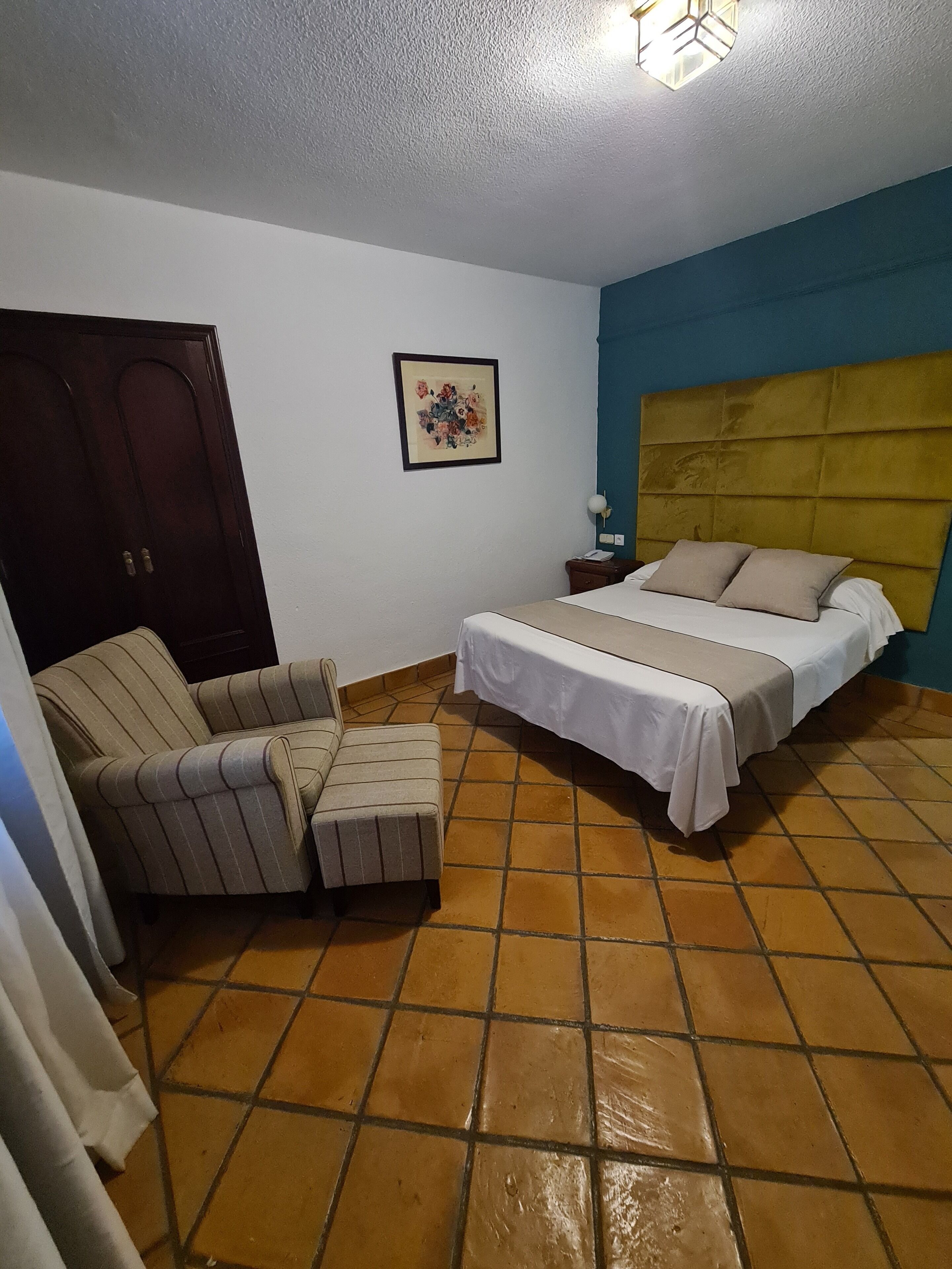 Quarto com cama, cadeira, mesa de cabeceira, luminária na parede e uma porta.