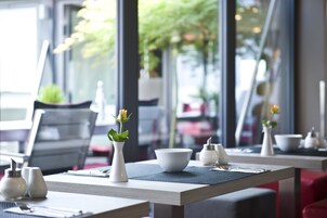 Interior de restaurante com mesas postas para refeição, um vaso com uma flor e vista para o exterior através de grandes janelas.